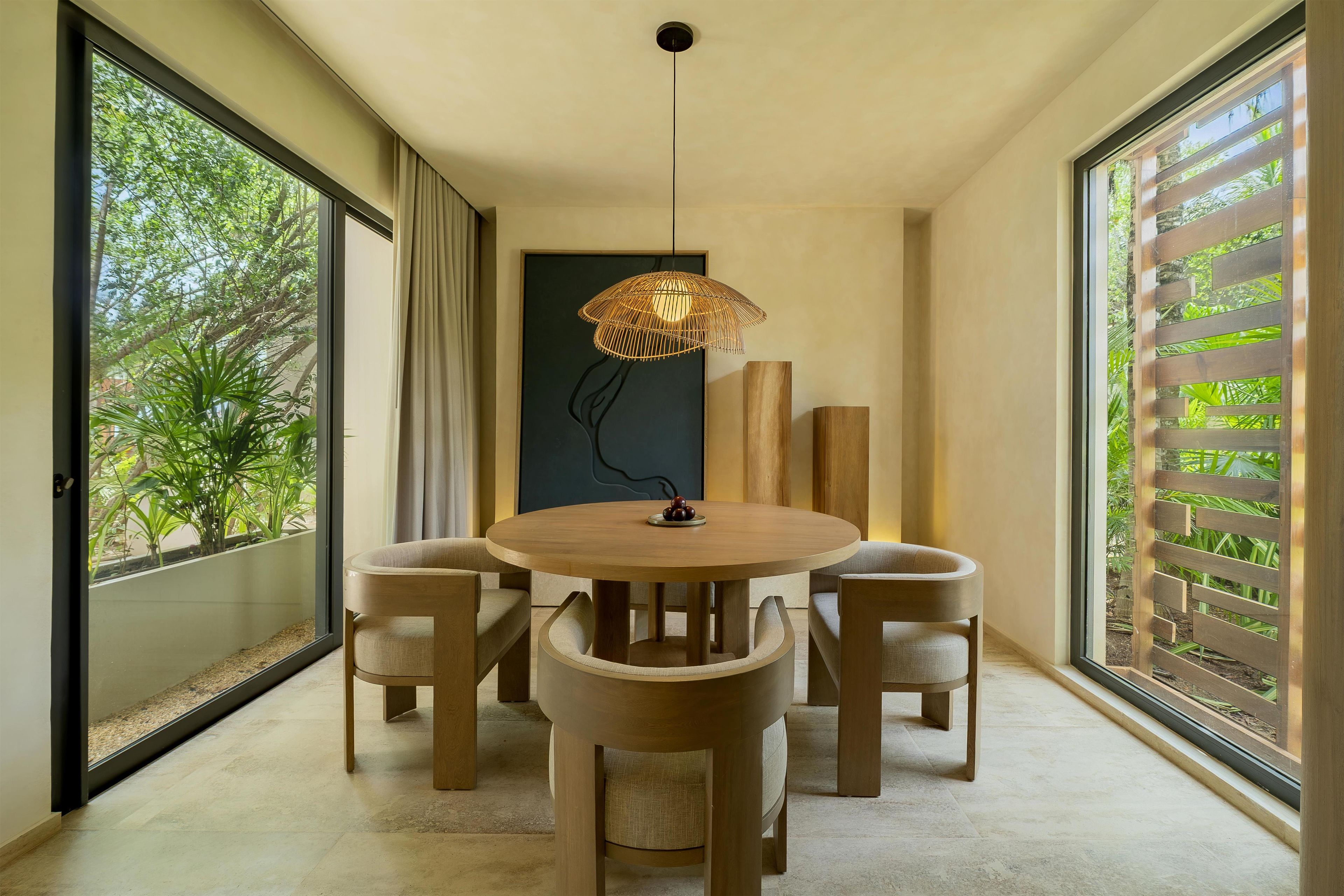 Uma sala de jantar com uma mesa redonda, quatro cadeiras, um lustre e vista para o verde através de grandes janelas.