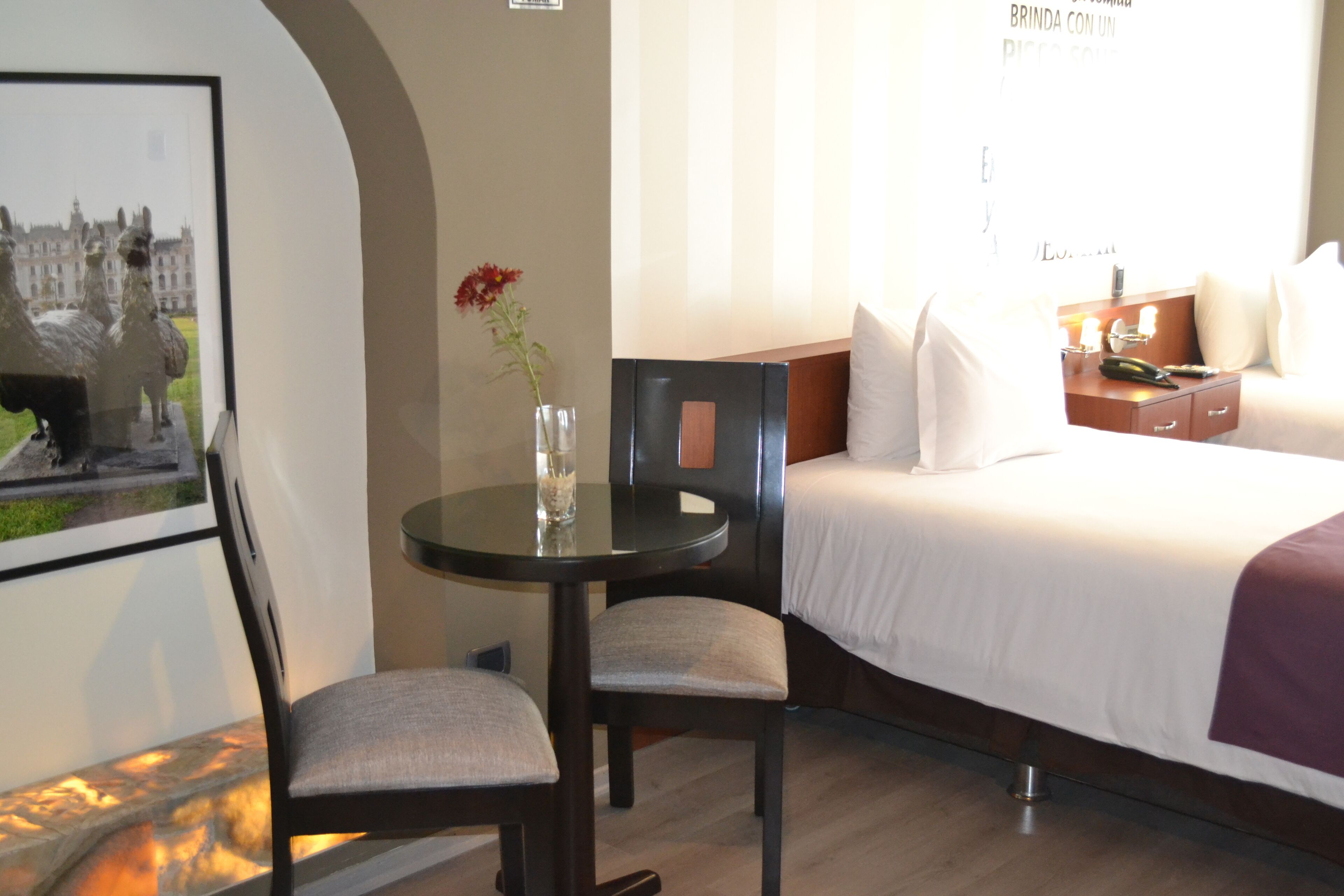 Quarto de hotel com uma cama, uma mesinha com um vaso de flores, uma cadeira e um quadro na parede.