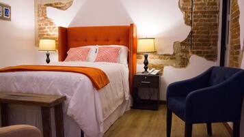 Quarto com parede de tijolos, cama de madeira, duas mesas de cabeceira com luminárias e uma cadeira azul.