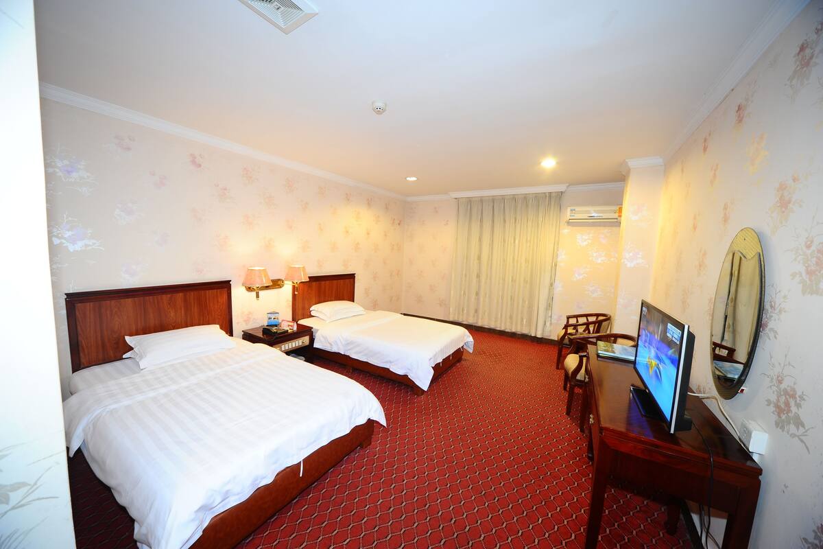 Quarto de hotel com duas camas, uma escrivaninha com computador, um espelho e uma janela com cortinas.