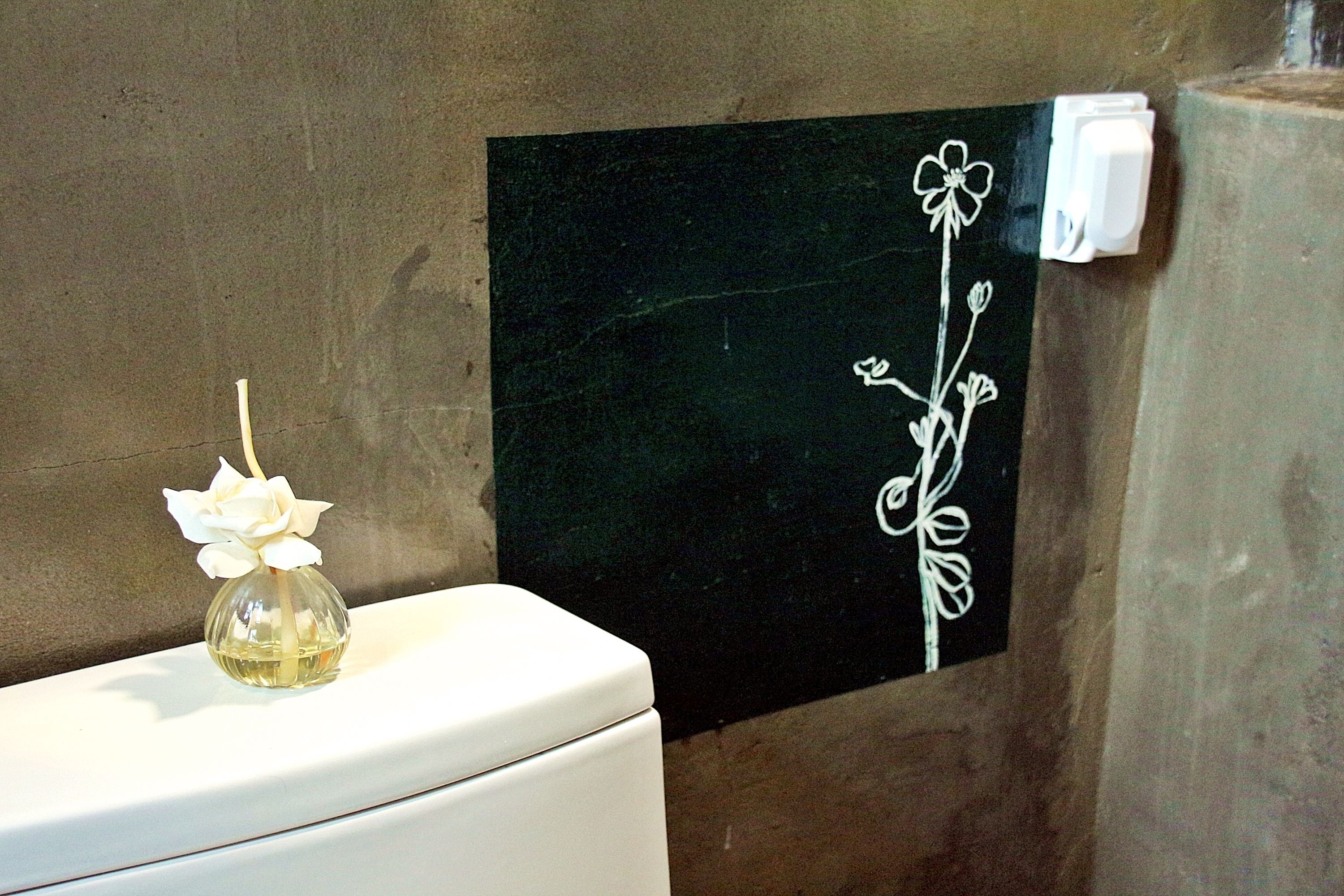 Uma casa de banho com sanita branca, um pequeno vaso com flores brancas e uma parede preta com um desenho floral branco.