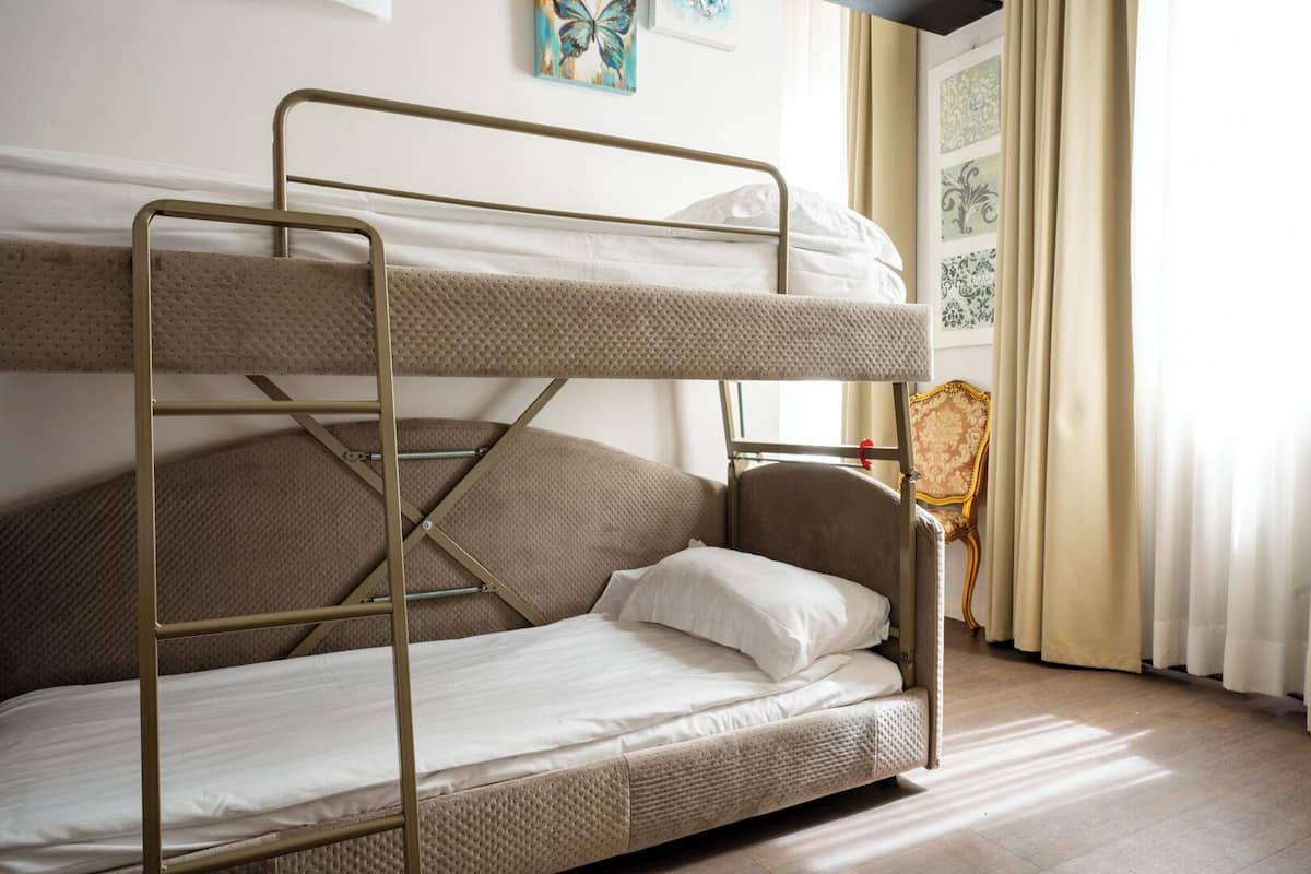 Uma cama de beliche com colchão e almofadas, uma cadeira e uma janela com cortinas.
