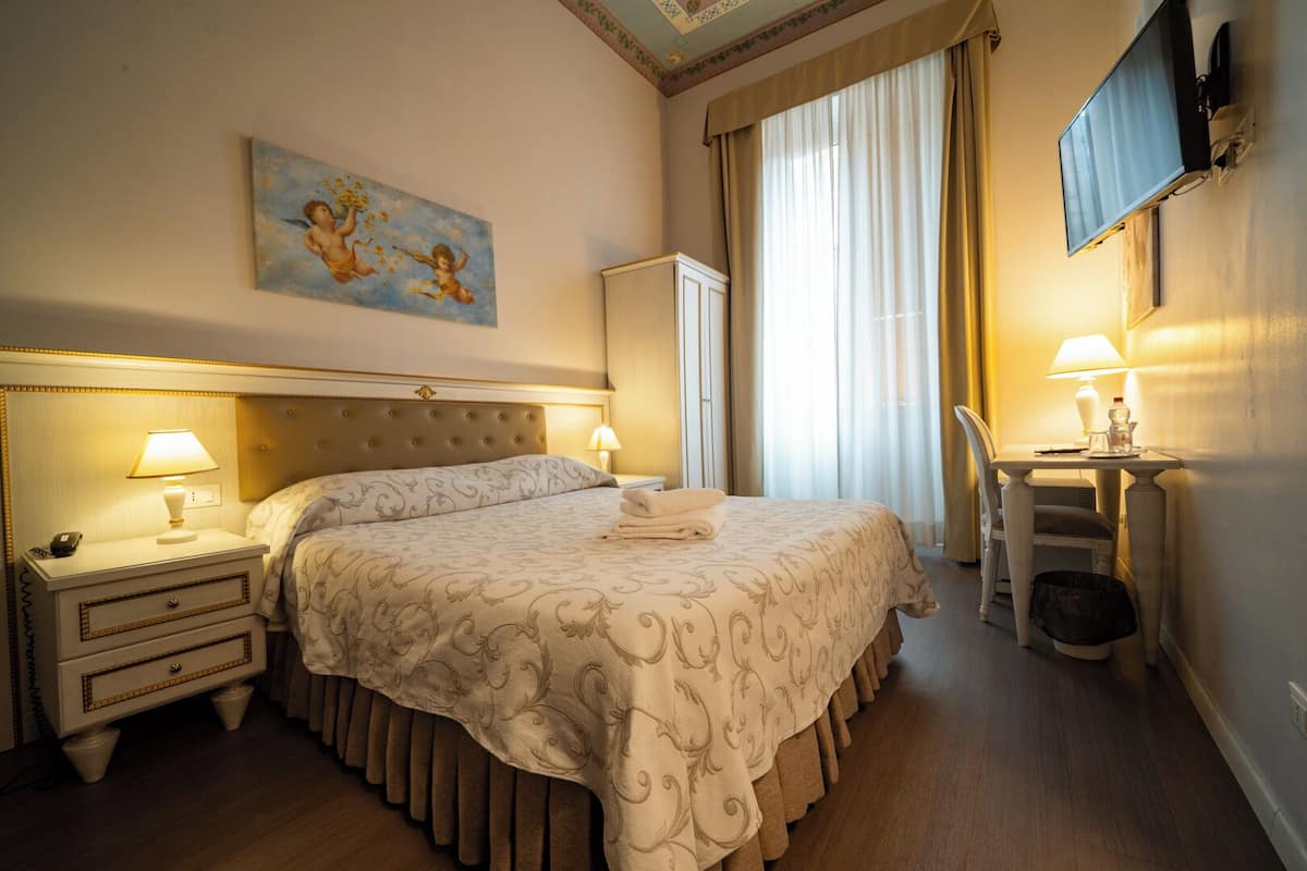 Quarto de hotel com uma cama grande, mesas de cabeceira com luminárias, uma escrivaninha com cadeira, uma televisão e um quadro na parede.
