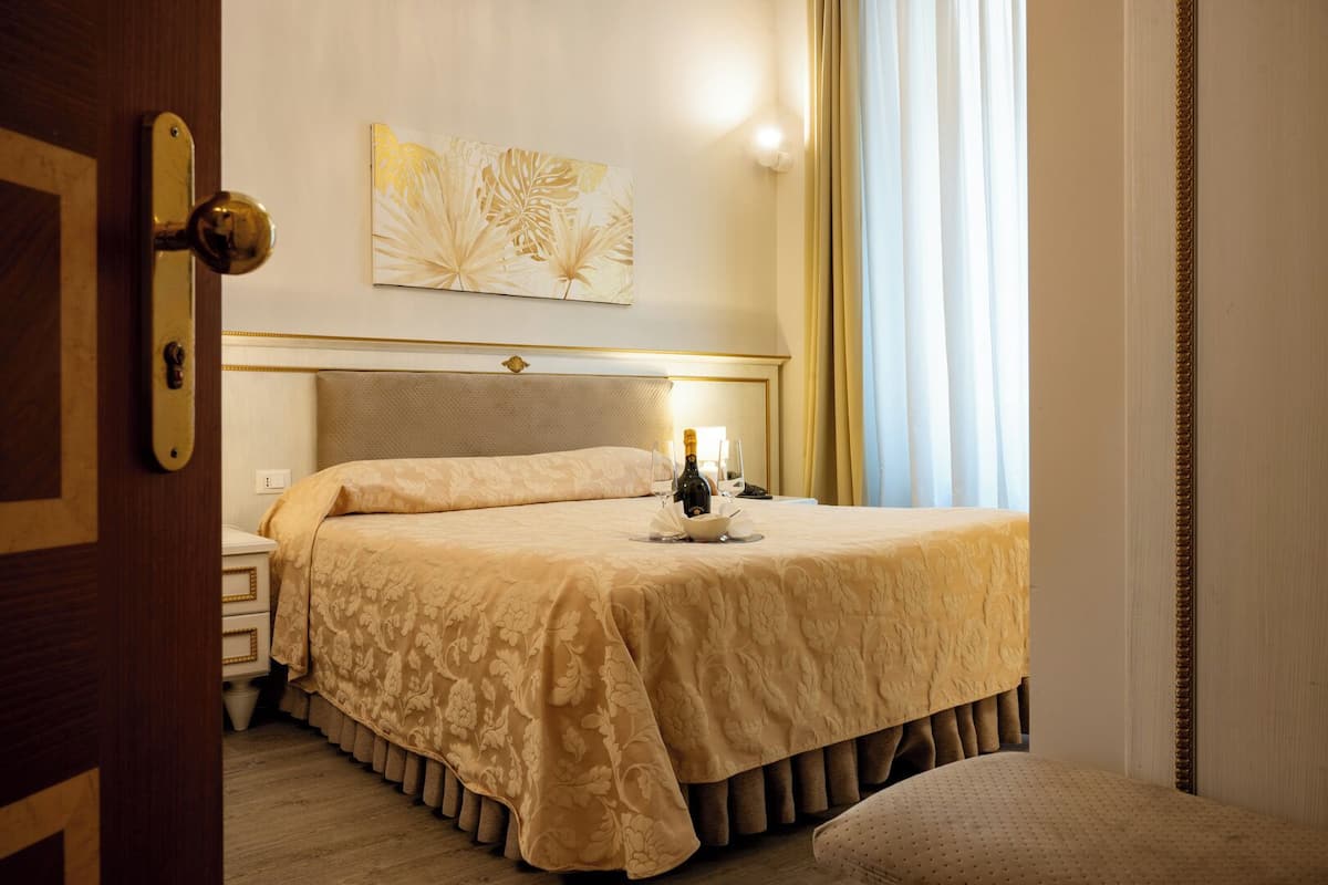 Quarto de hotel com cama, mesa de cabeceira, uma garrafa de champanhe e uma obra de arte na parede.