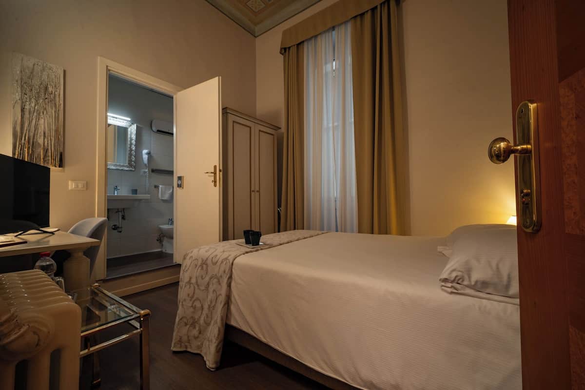 Quarto de hotel com cama, escrivaninha com cadeira, banheiro com chuveiro e guarda-roupa.