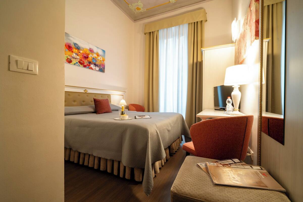 Quarto de hotel com uma cama, escrivaninha com luminária, cadeira, televisão e um quadro na parede.