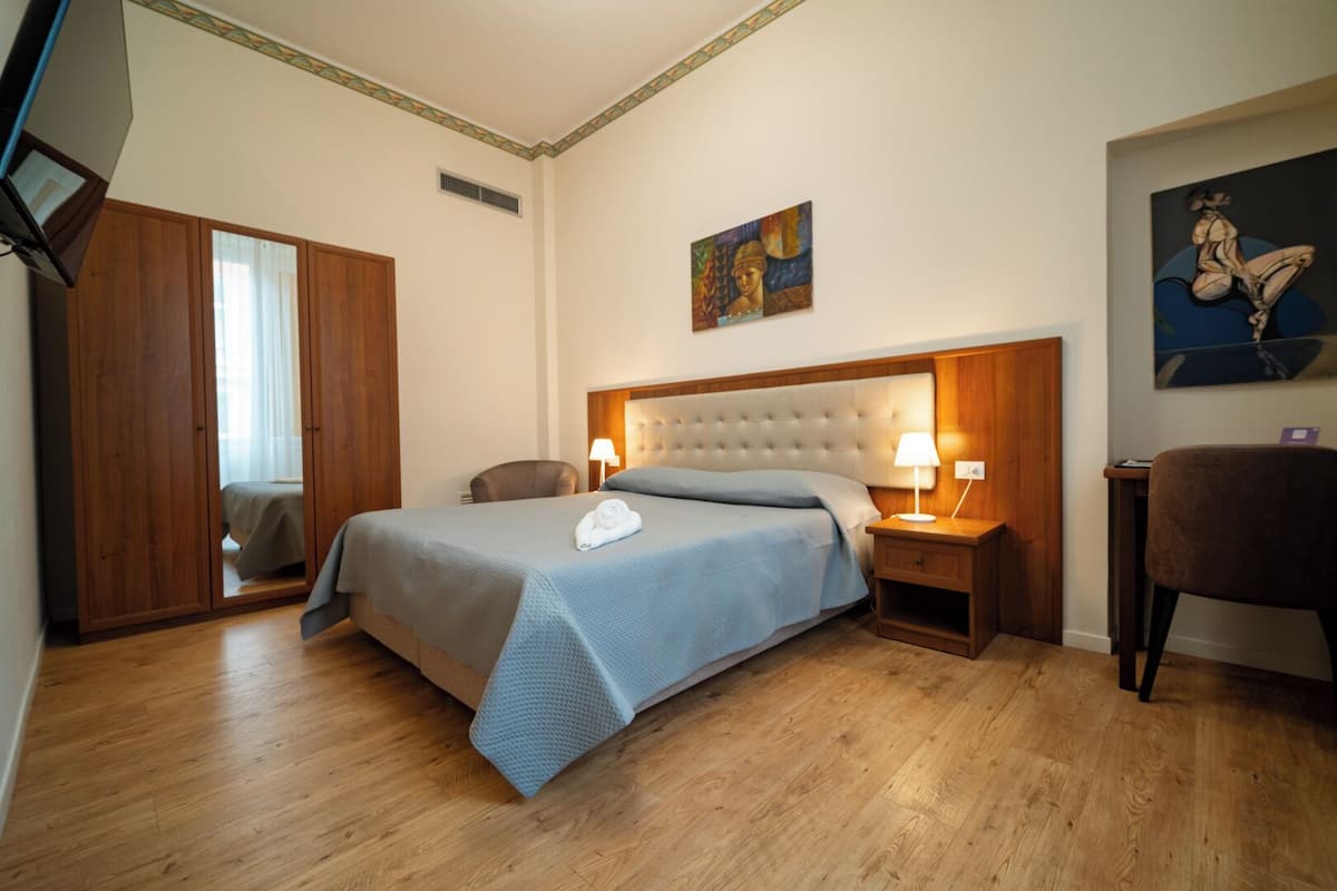 Quarto de hotel com cama, mesa de cabeceira, cadeira, guarda-roupa de madeira e obras de arte nas paredes.