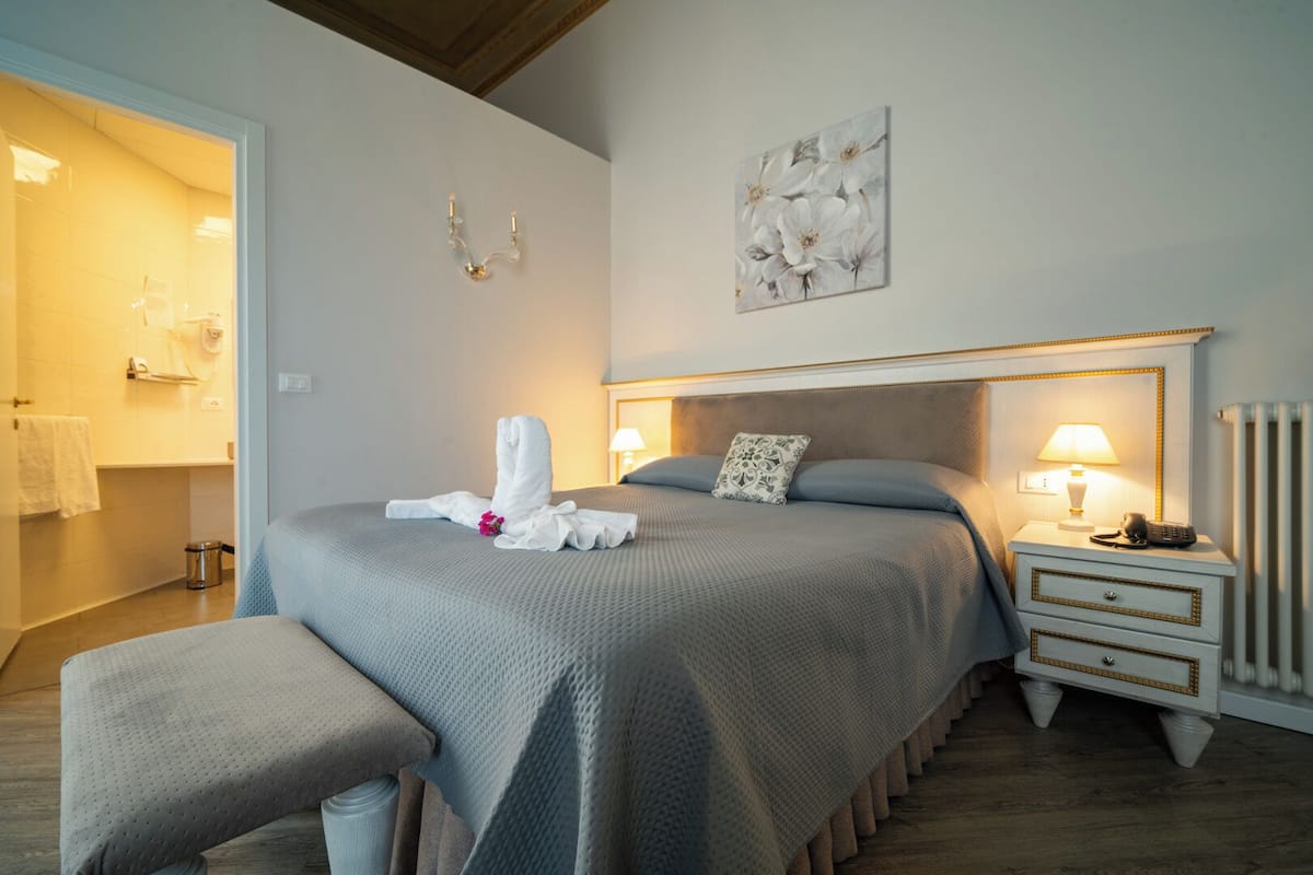 Quarto de hotel com cama, mesas de cabeceira, um banco, luminária na parede e um quadro.