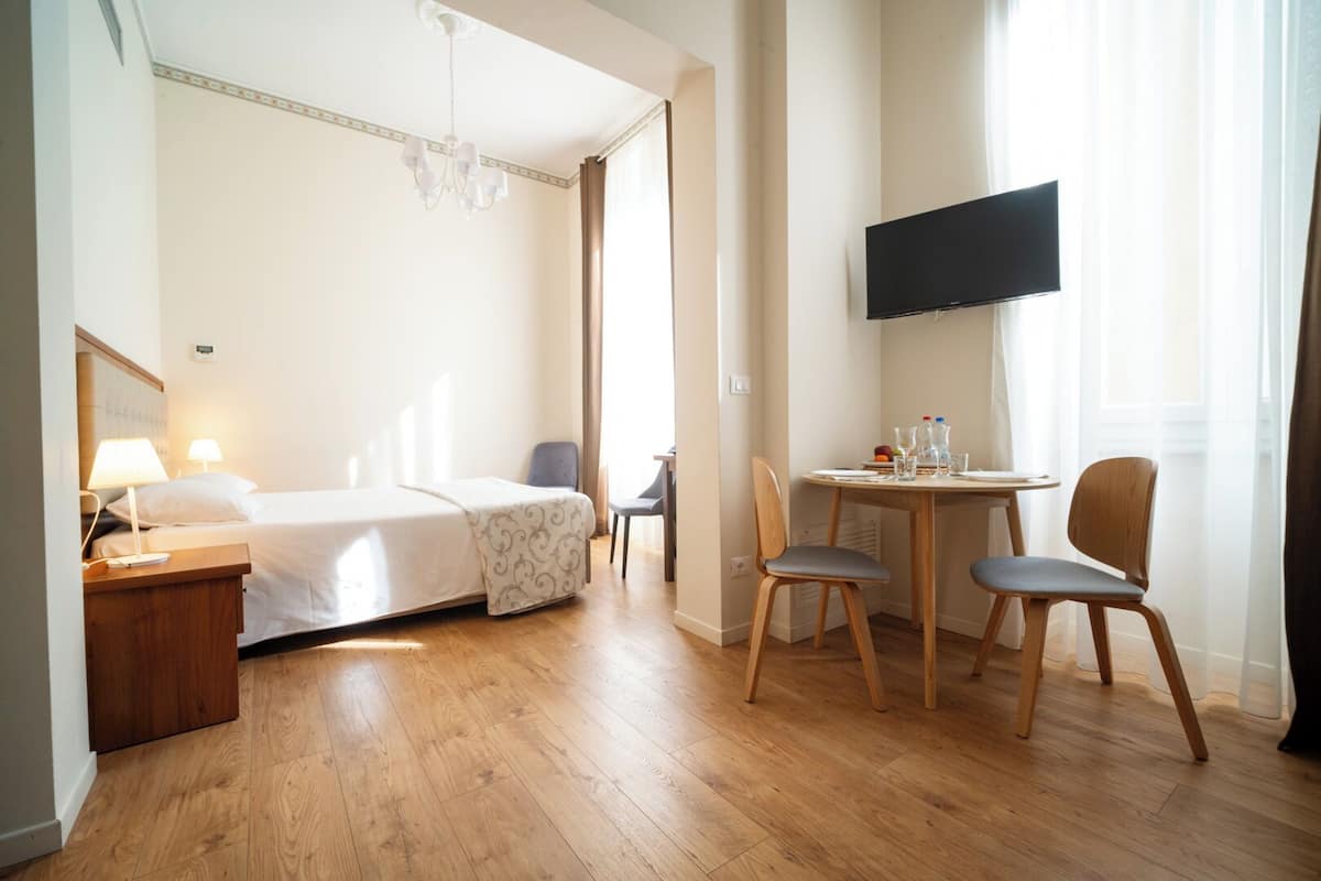 Quarto de hotel com cama, mesa de cabeceira, luminária, televisão e área de refeições com cadeiras e uma mesa.