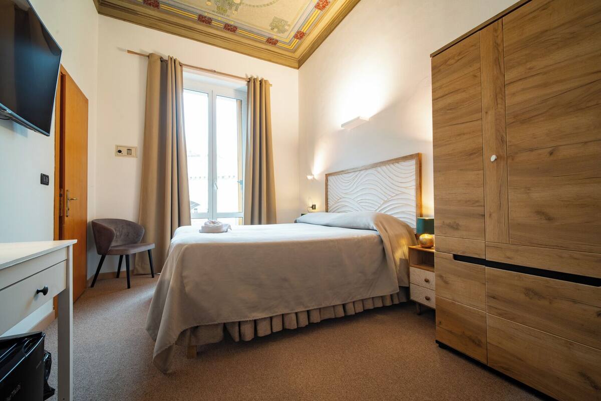 Quarto de hotel com cama, escrivaninha, cadeira, guarda-roupa e televisão.