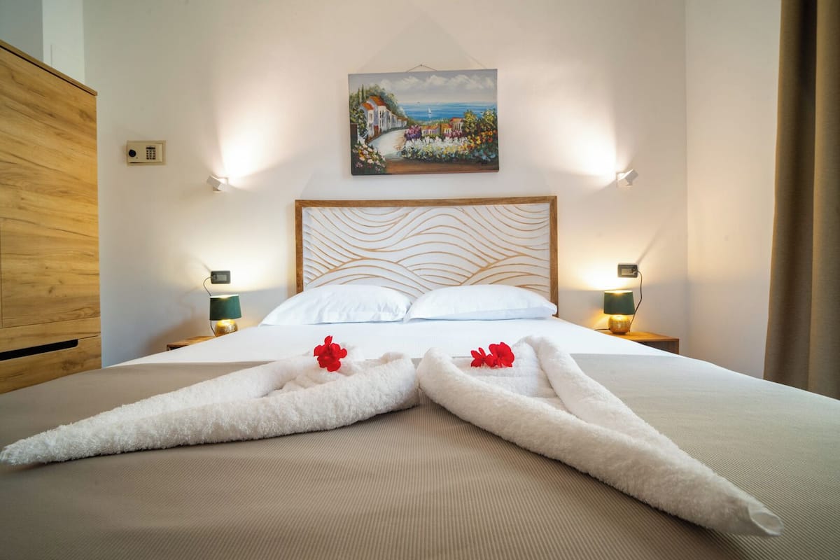 Quarto de hotel com uma cama, duas mesas de cabeceira, um quadro na parede e um guarda-roupa de madeira.
