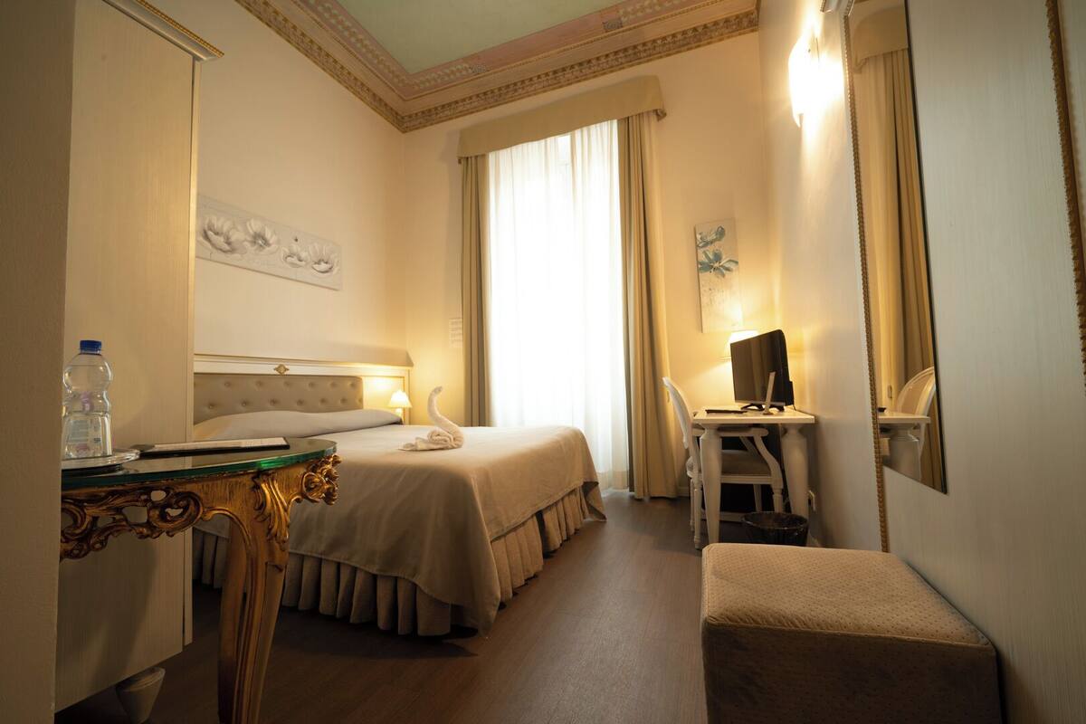 Quarto de hotel com cama, escrivaninha com computador, cadeira, mesa de cabeceira com garrafa de água e espelho.