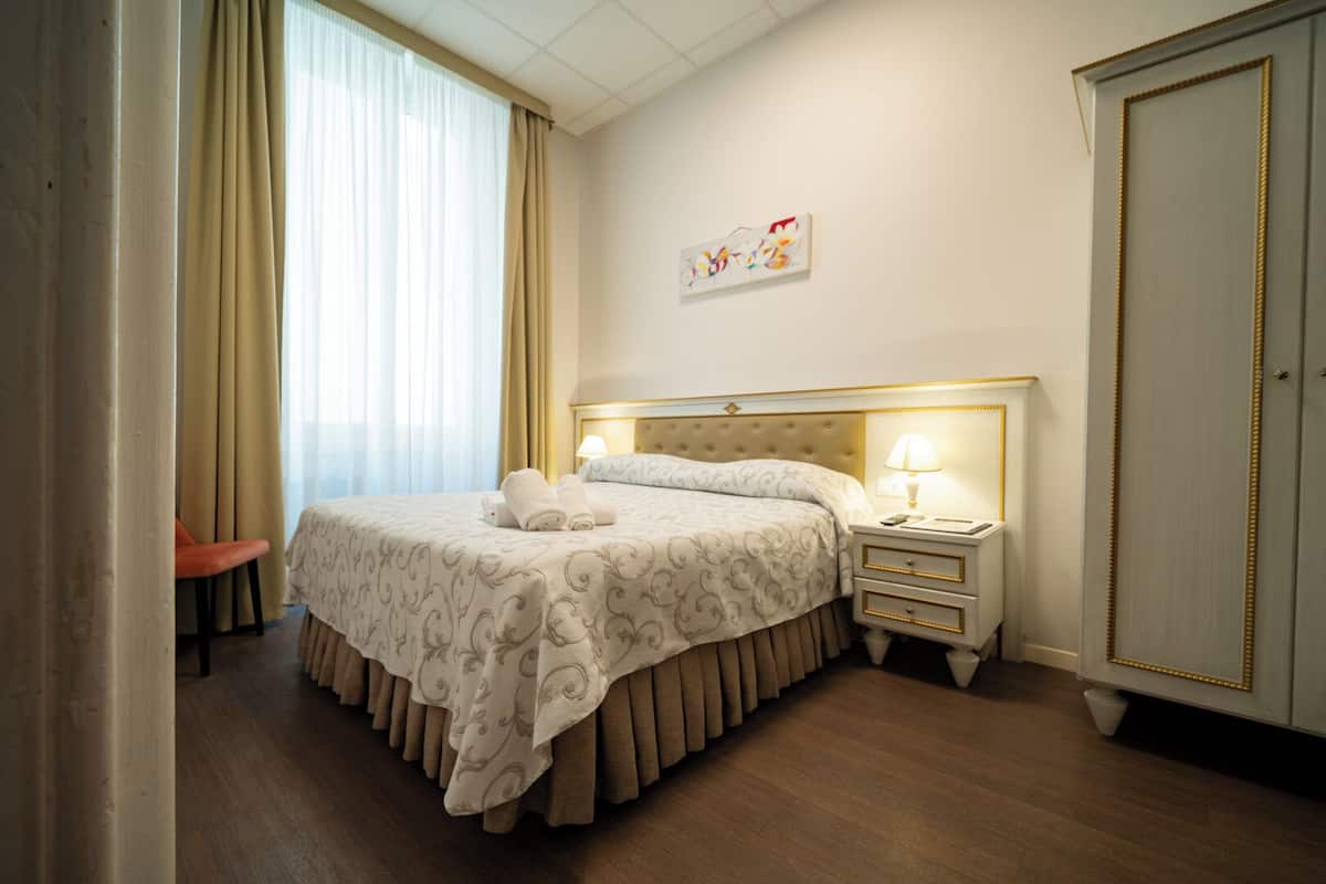 Quarto de hotel com cama, mesas de cabeceira, armário e janela com cortinas.