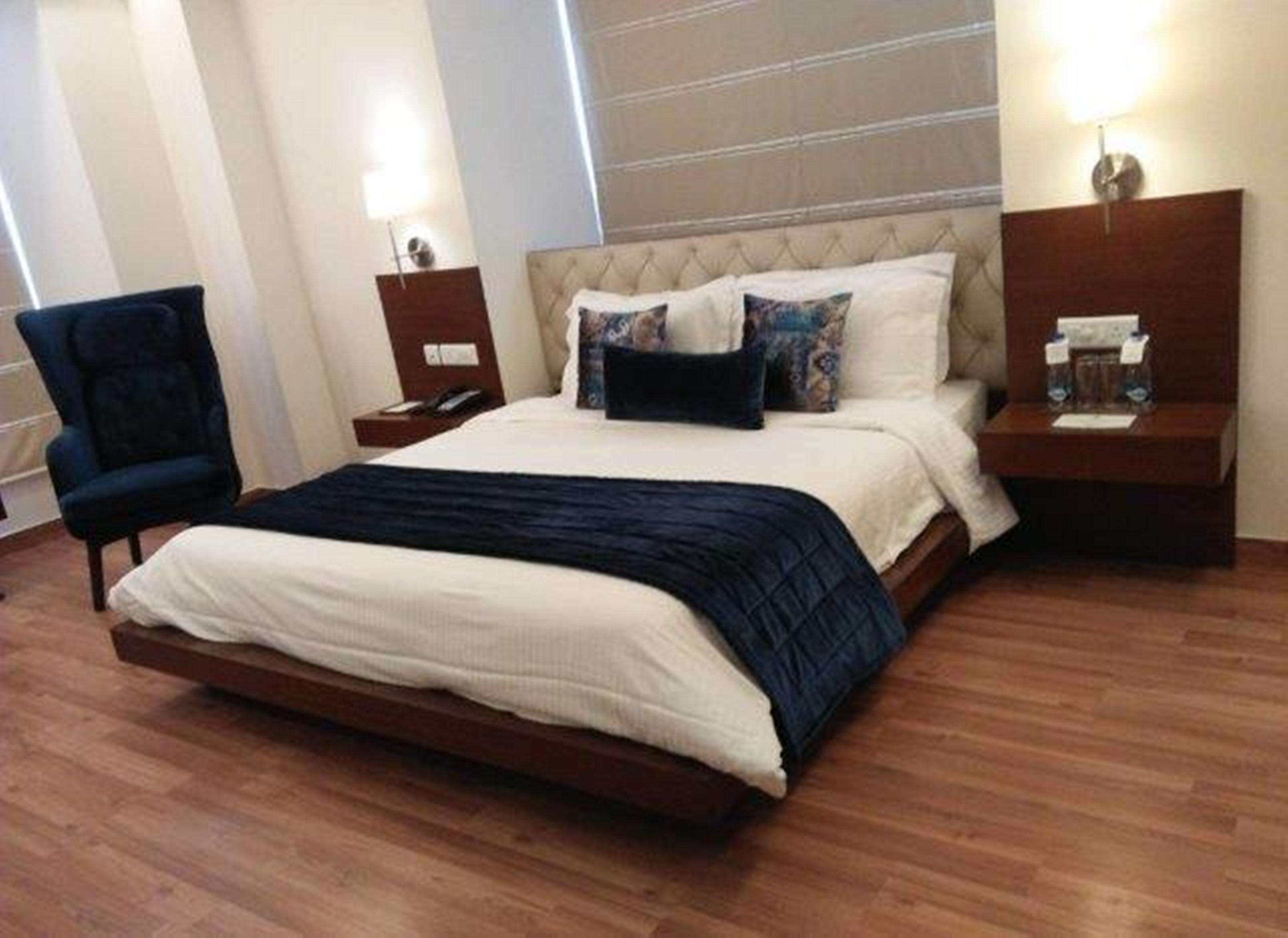 Quarto de hotel com uma cama grande, uma poltrona, uma mesinha de cabeceira e um criado-mudo.