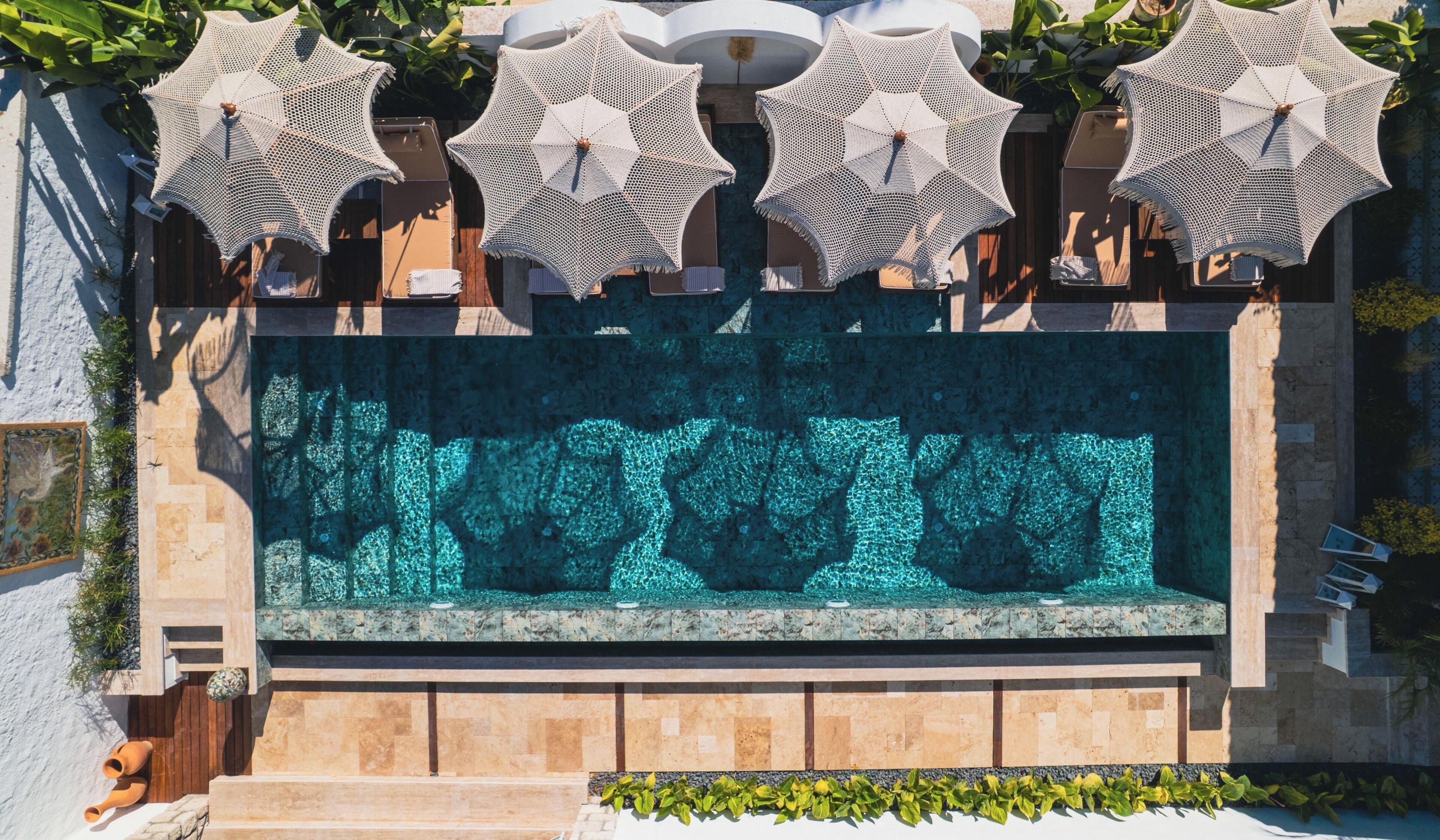 Uma área de piscina no terraço com vários guarda-sóis brancos, uma parede de pedra e uma piscina azul límpida.