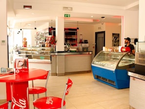 Um café com mesas e cadeiras com a marca Coca-Cola em vermelho, uma vitrine de vidro com alimentos e uma pessoa trabalhando atrás do balcão.