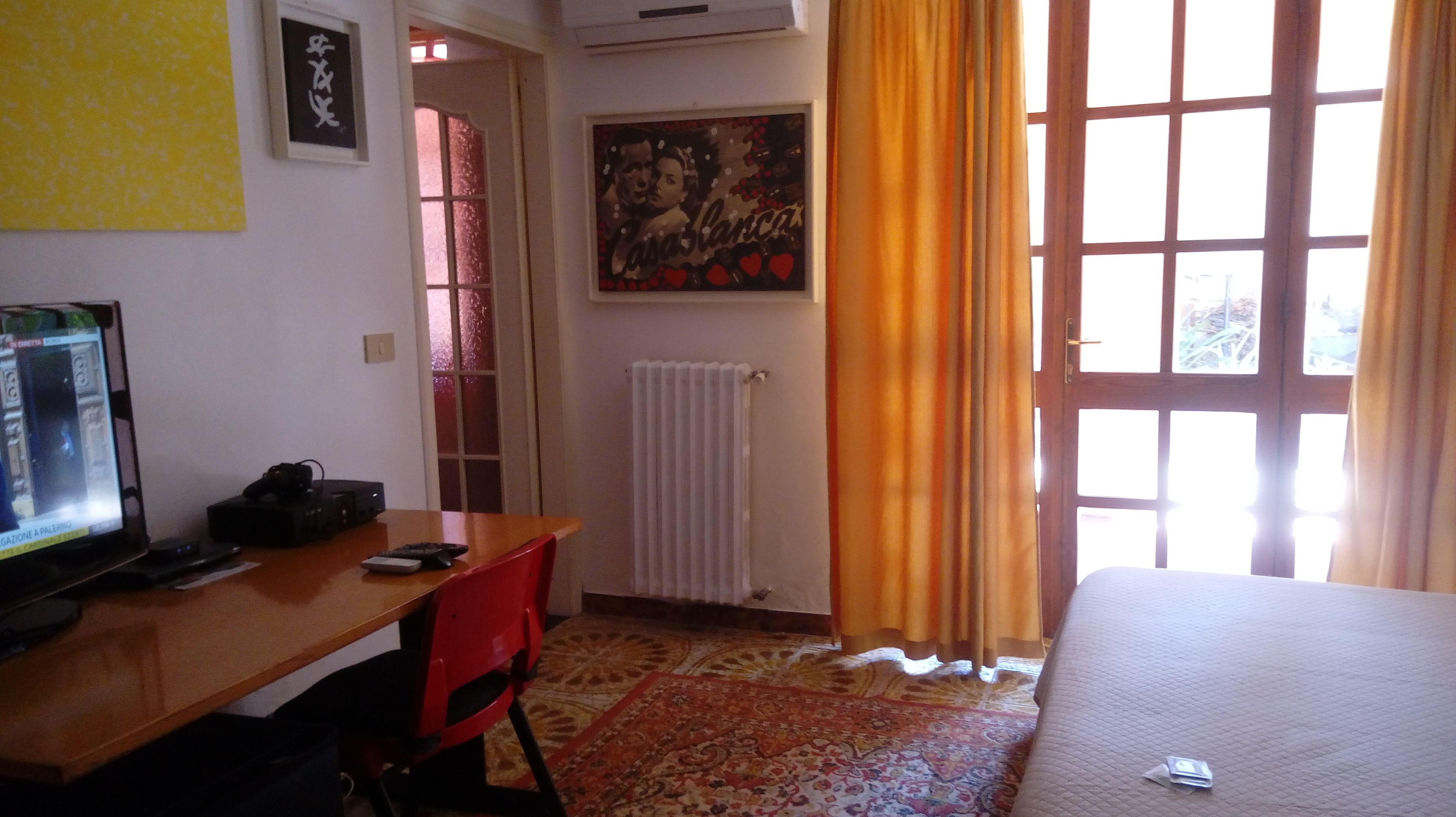 Quarto de hotel com secretária de madeira, uma cadeira vermelha, cama, radiador e janela com cortinas.