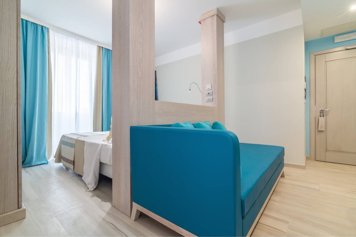 Quarto moderno com cama, sofá azul, coluna de madeira e uma porta.