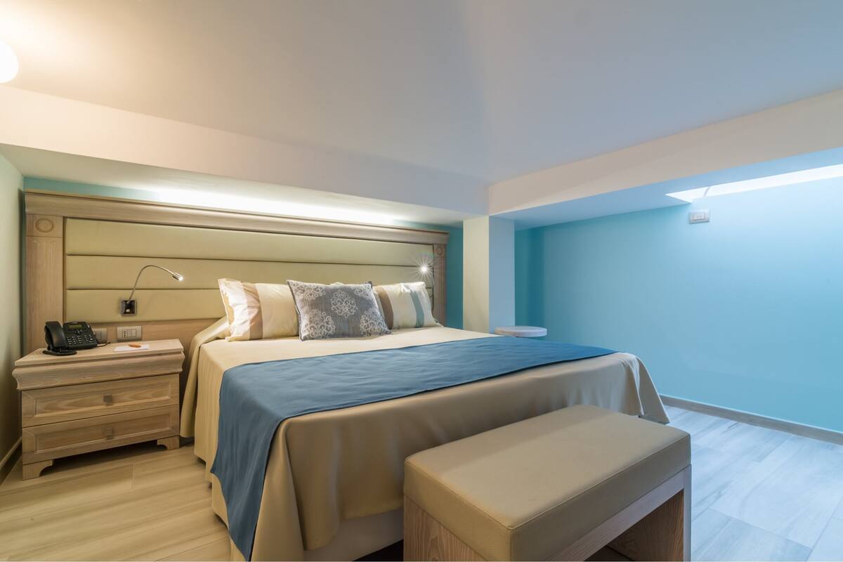 Quarto com cama, mesas de cabeceira, um banquinho e luminária fixada na parede.