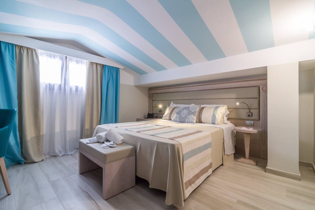 Quarto de hotel com cama, mesa de cabeceira, bancos, cortinas e teto com padrões listrados.