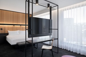 Quarto de hotel moderno com uma cama, uma escrivaninha, uma cadeira, uma televisão e uma janela grande com cortinas.