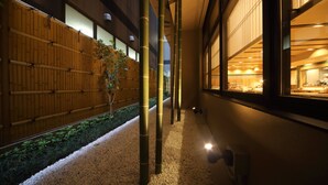 Um corredor com cerca de bambu, pedregulhos e vista para uma área de restaurante.