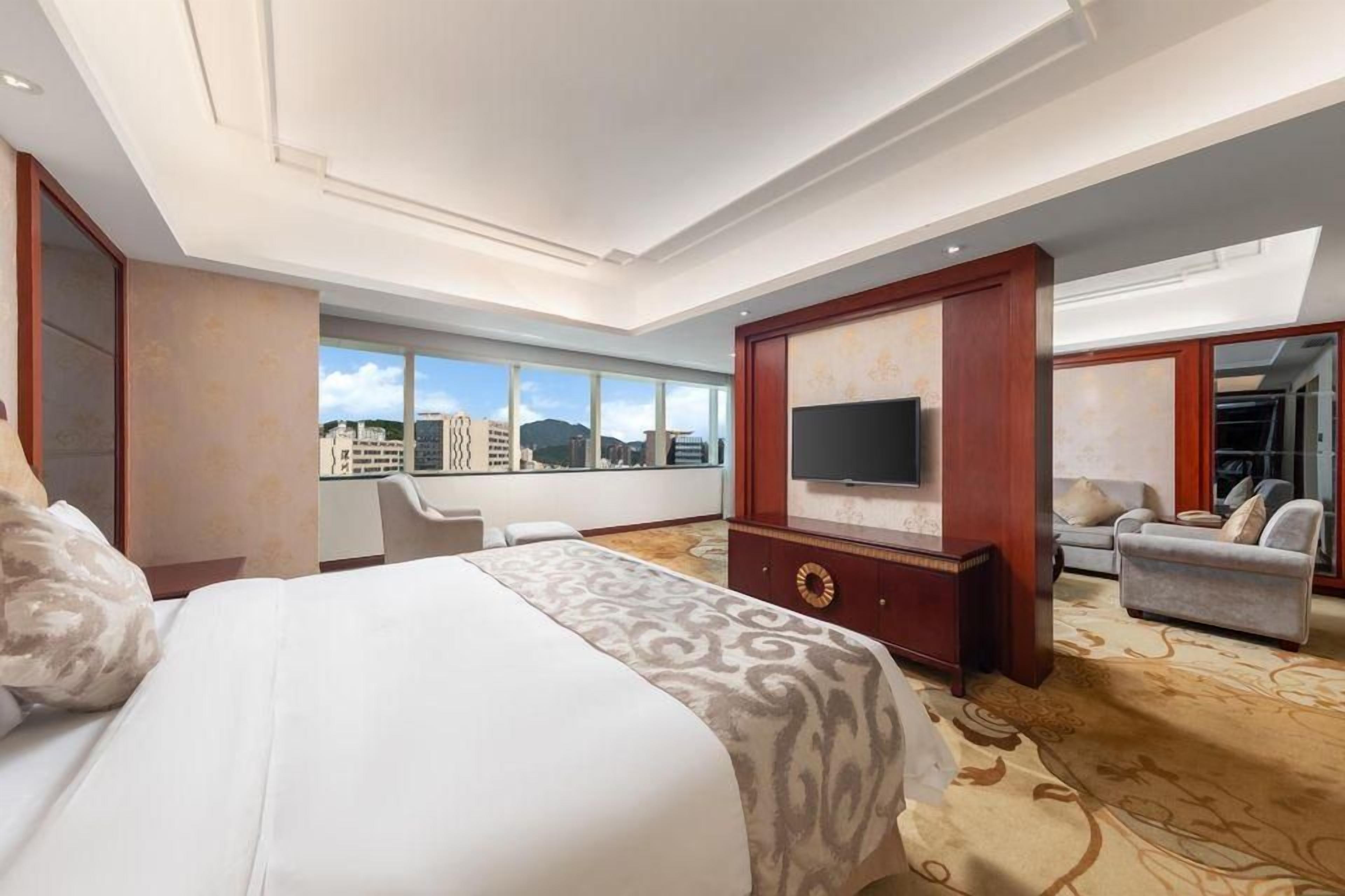 Quarto de hotel com uma cama grande, televisão, sofá e vista para a paisagem urbana.