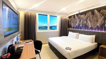 Quarto de hotel com cama, escrivaninha com cadeira, televisão e vista para o mar através de duas janelas.