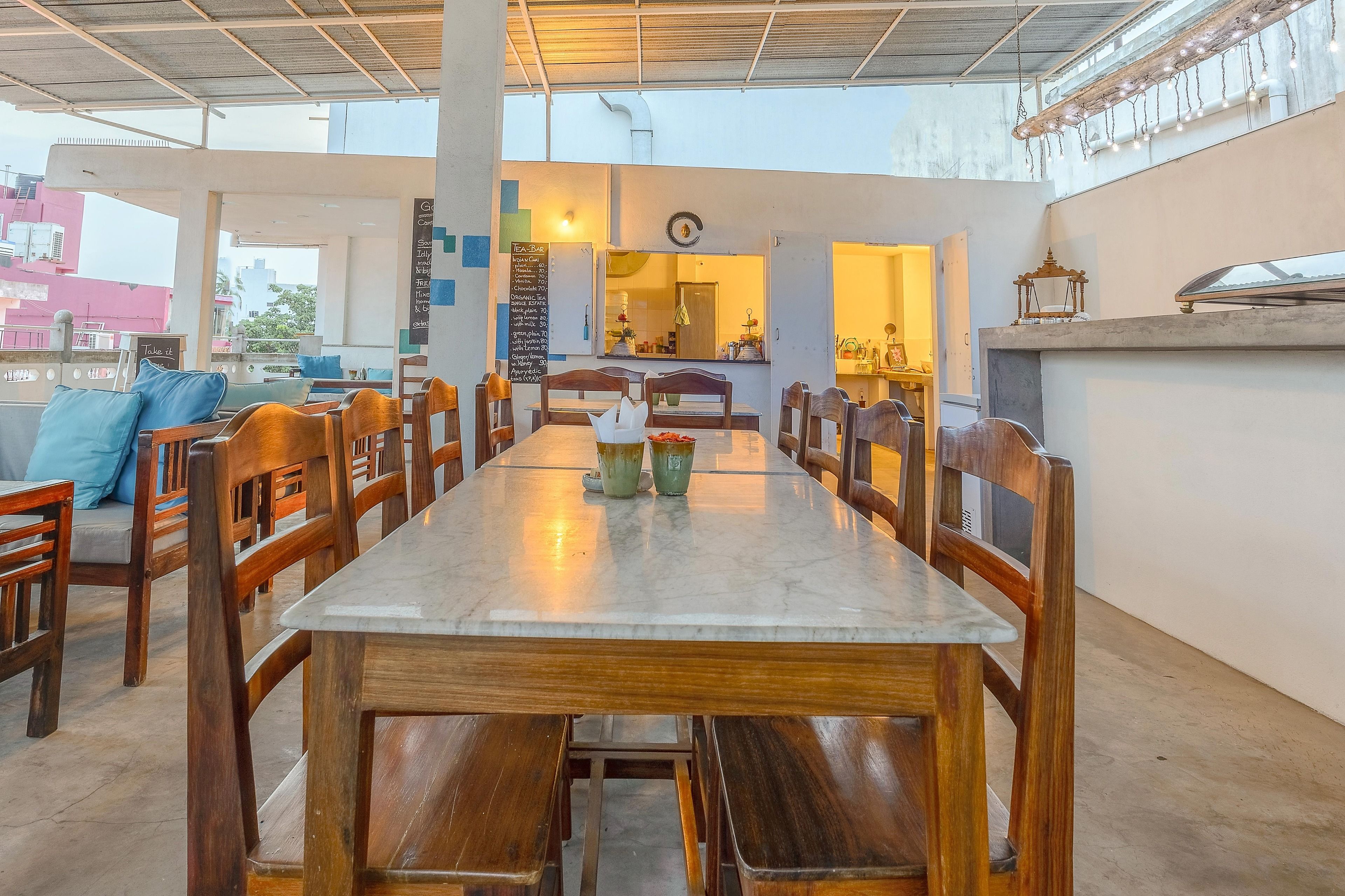 Uma ampla área de refeições ao ar livre, com uma mesa comprida e cadeiras de madeira, um espaço de estar pequeno com almofadas e vista para a cidade ao fundo.