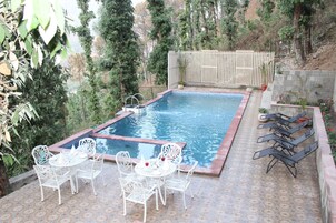 Piscina com deck ladrilhado, rodeada por cadeiras e mesa brancas, com cerca e vegetação ao fundo.