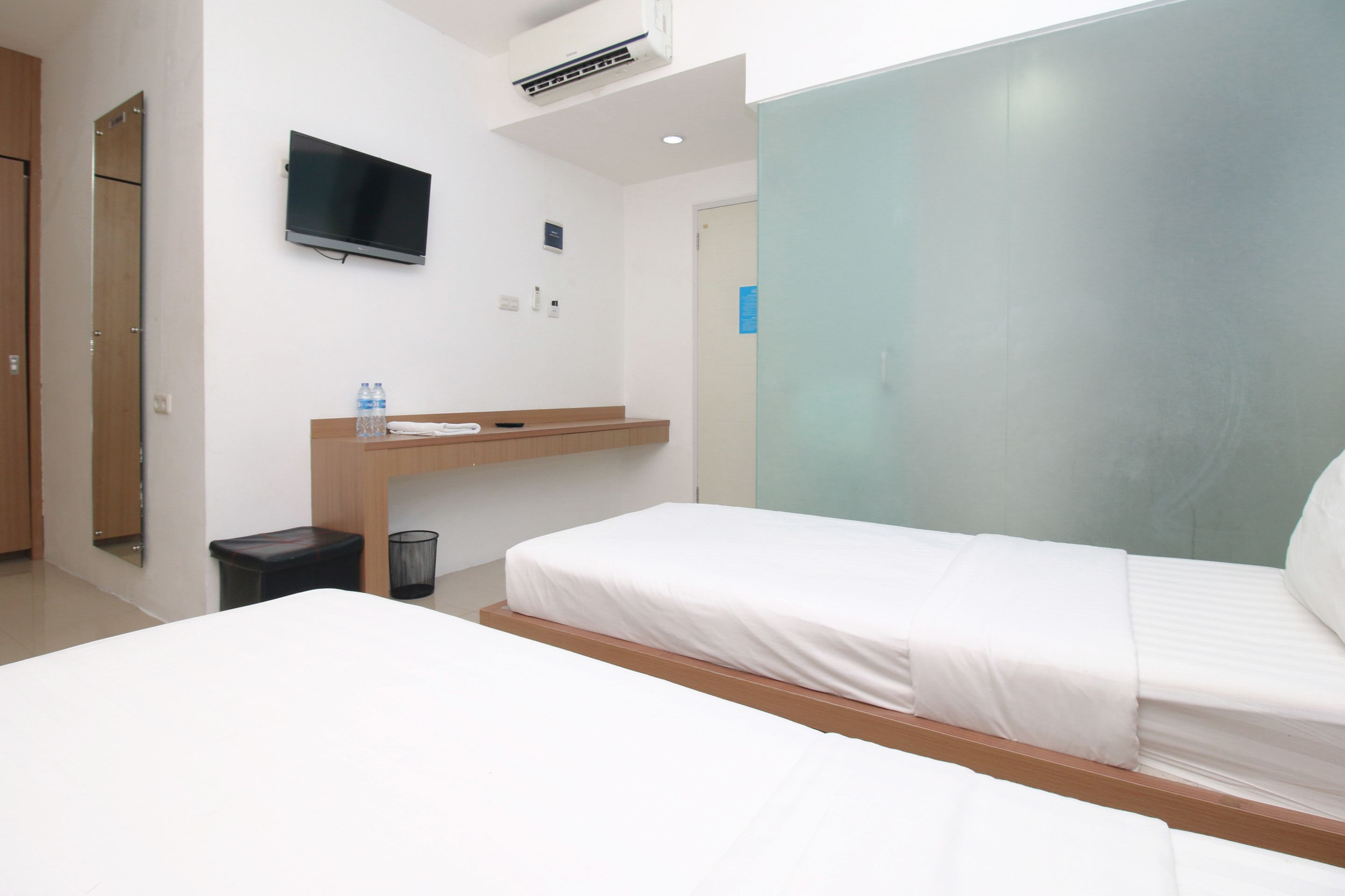 Quarto de hotel com uma cama, televisão na parede, uma mesinha com uma garrafa de água e uma lixeira.