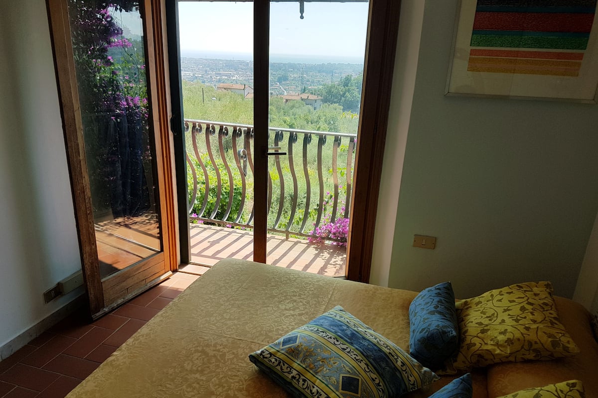Quarto com uma cama grande, duas almofadas e vista para uma varanda com grade e vegetação.