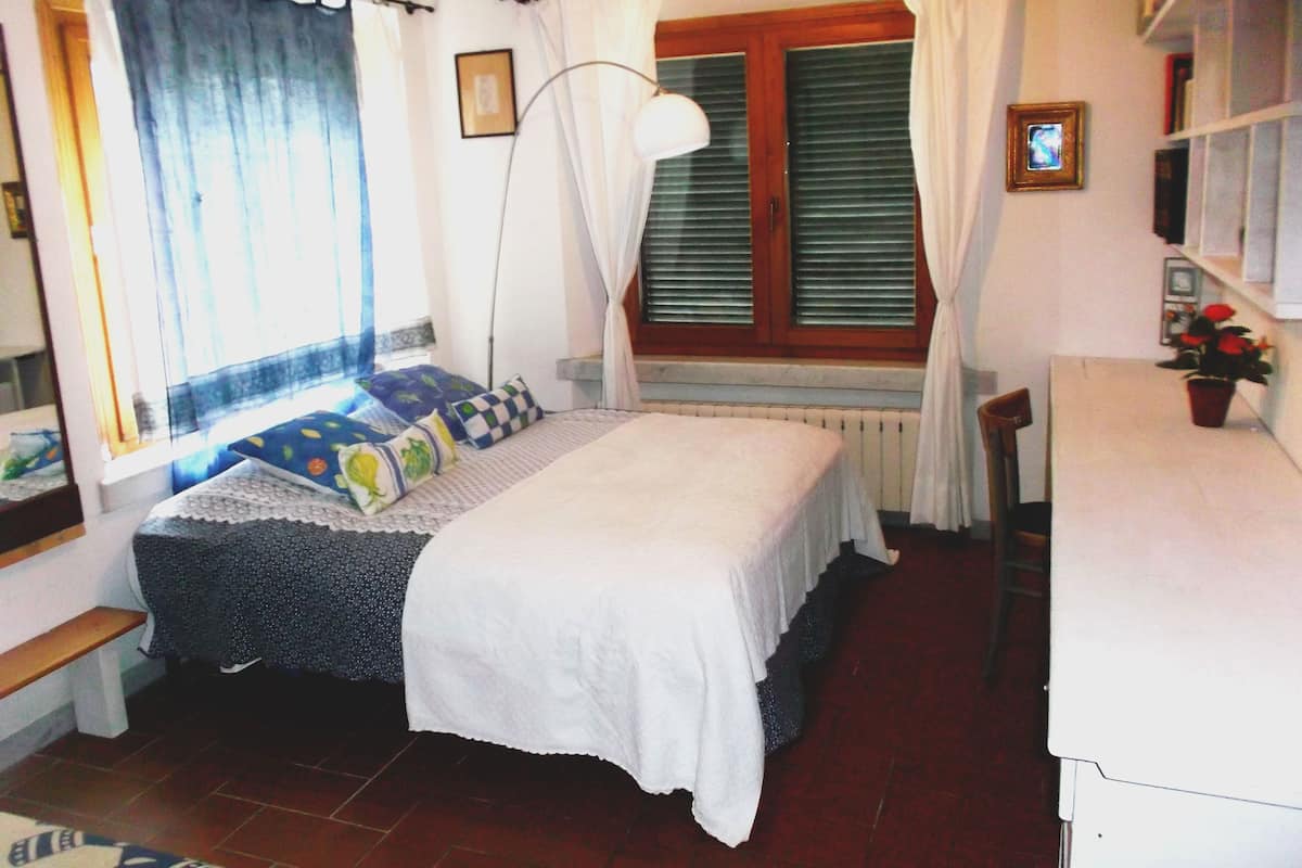 Um quarto com cama, janela com cortinas, radiador, escrivaninha e uma cadeira.