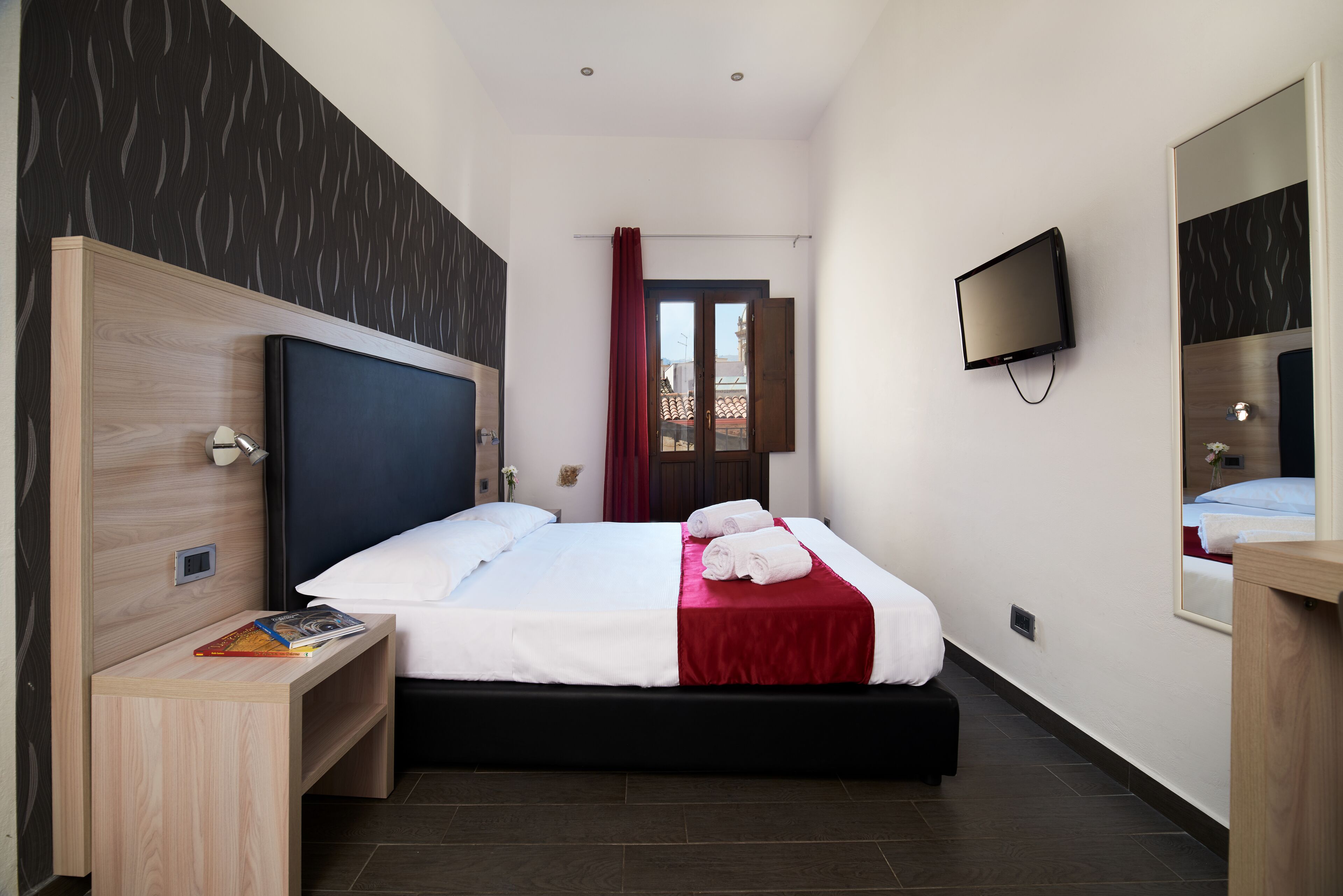 Quarto de hotel moderno com uma cama grande, uma mesinha de cabeceira com um livro, uma TV na parede e um espelho sobre a cama.