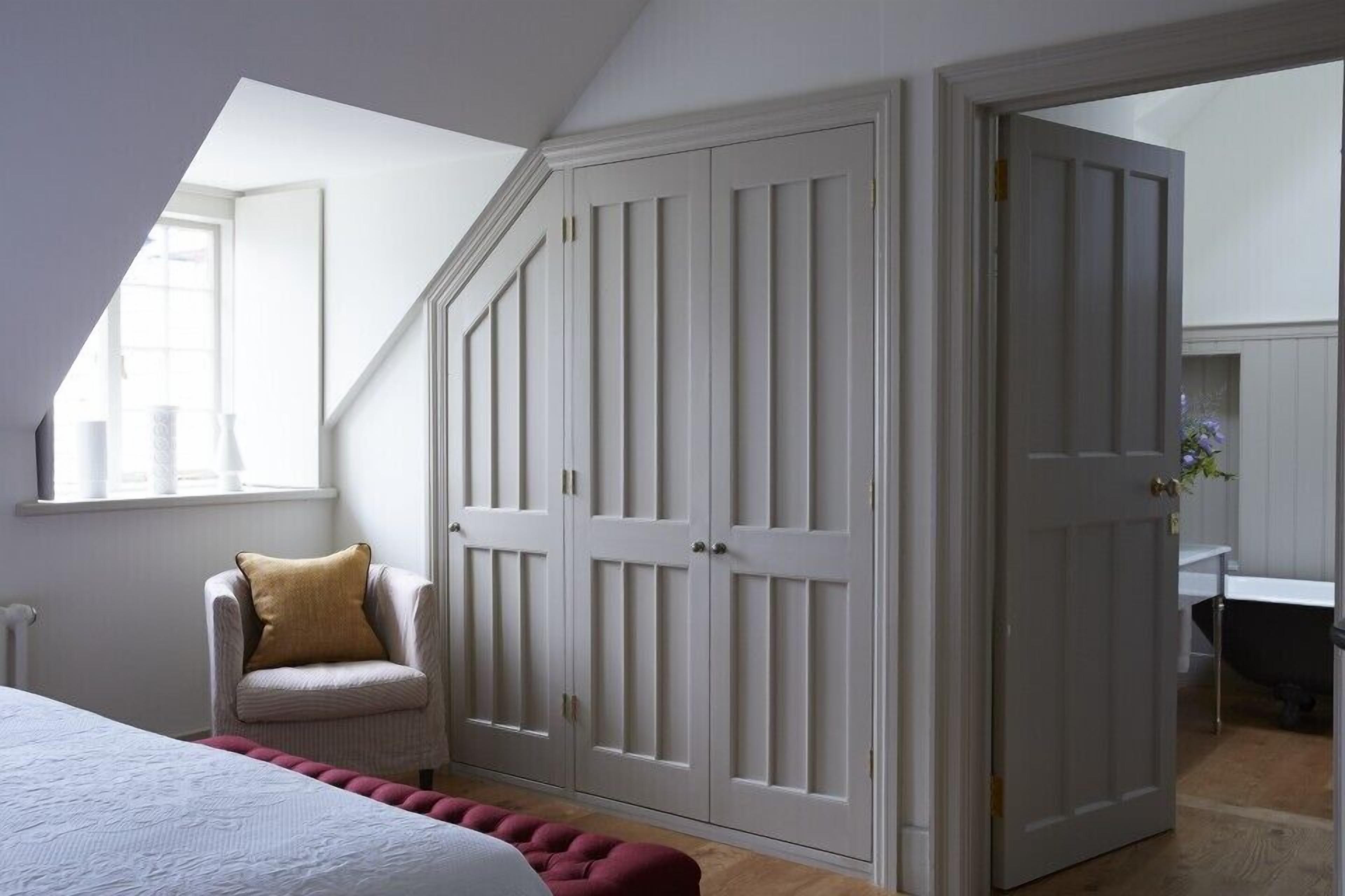 Quarto com teto inclinado, uma cama, uma cadeira, um guarda-roupa e um banheiro visível por uma porta aberta.