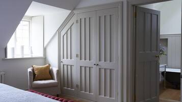 Quarto com teto inclinado, uma cama, uma cadeira, um guarda-roupa e um banheiro visível por uma porta aberta.