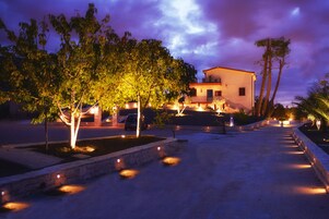 Edifício bem iluminado com calçada pavimentada e áreas ajardinadas, cercado por árvores e céu claro.