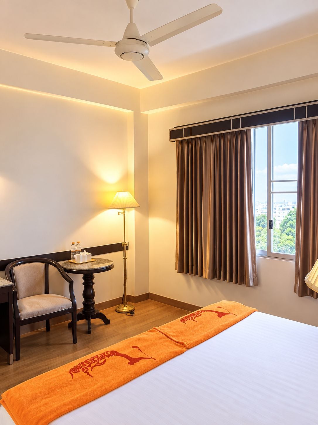 Quarto de hotel com cama, escrivaninha, cadeira, luminária de chão e janela com cortinas.