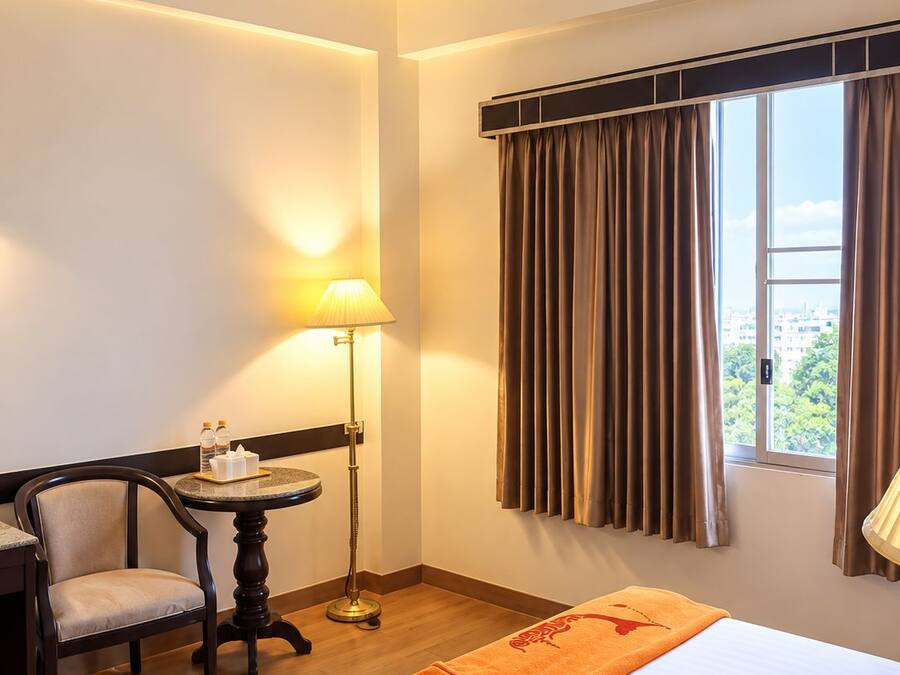Quarto de hotel com cama, escrivaninha, cadeira, luminária de chão e janela com cortinas.