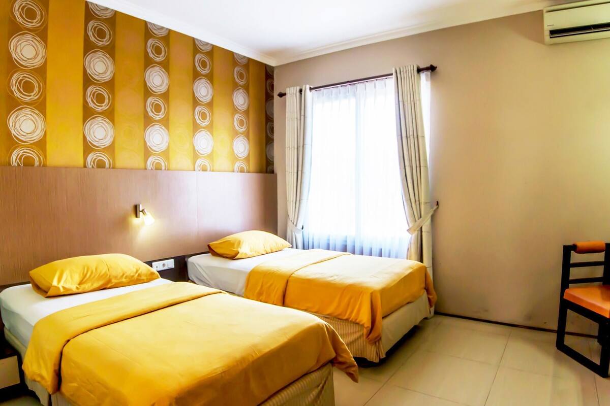 Quarto de hotel com duas camas, uma cadeira, uma janela com cortinas e parede com padrão circular.