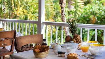 Mesa de café da manhã posta com croissants, frutas e bebidas, com vista para vegetação exuberante.