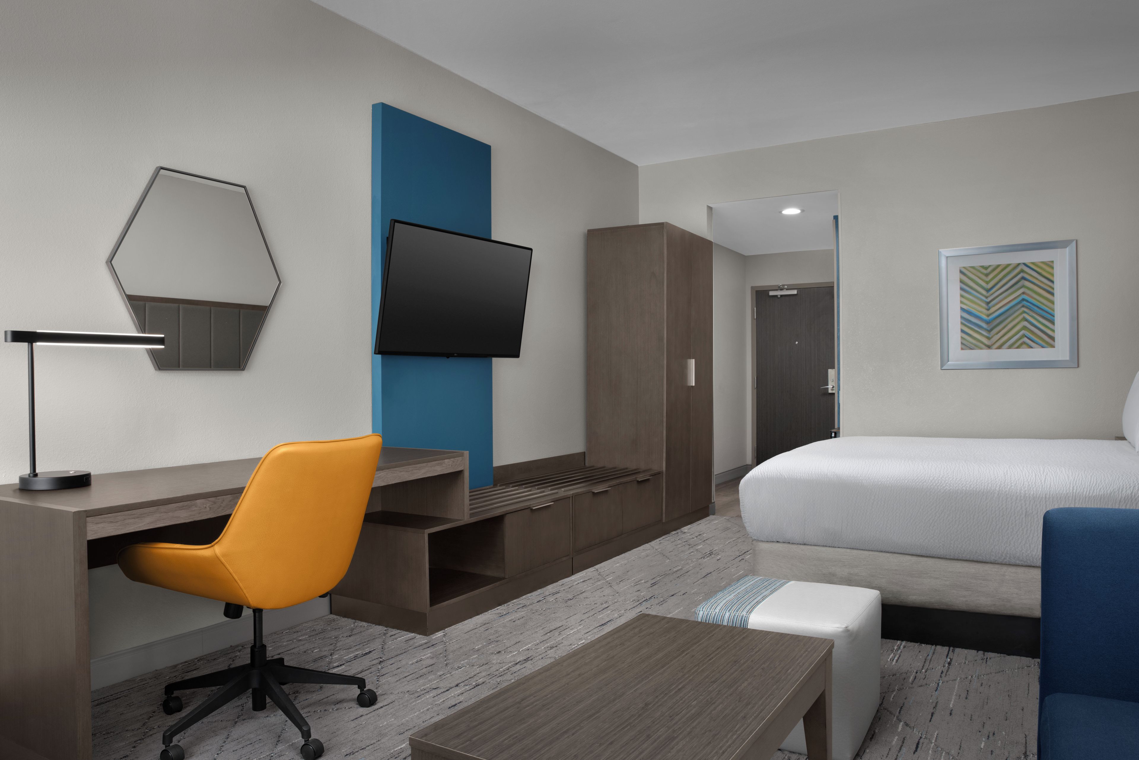 Quarto de hotel com cama, escrivaninha, cadeira, televisão e guarda-roupa.