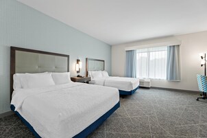 Quarto de hotel com duas camas, uma mesa de cabeceira, abajur, janela com cortinas e piso coberto por carpete.