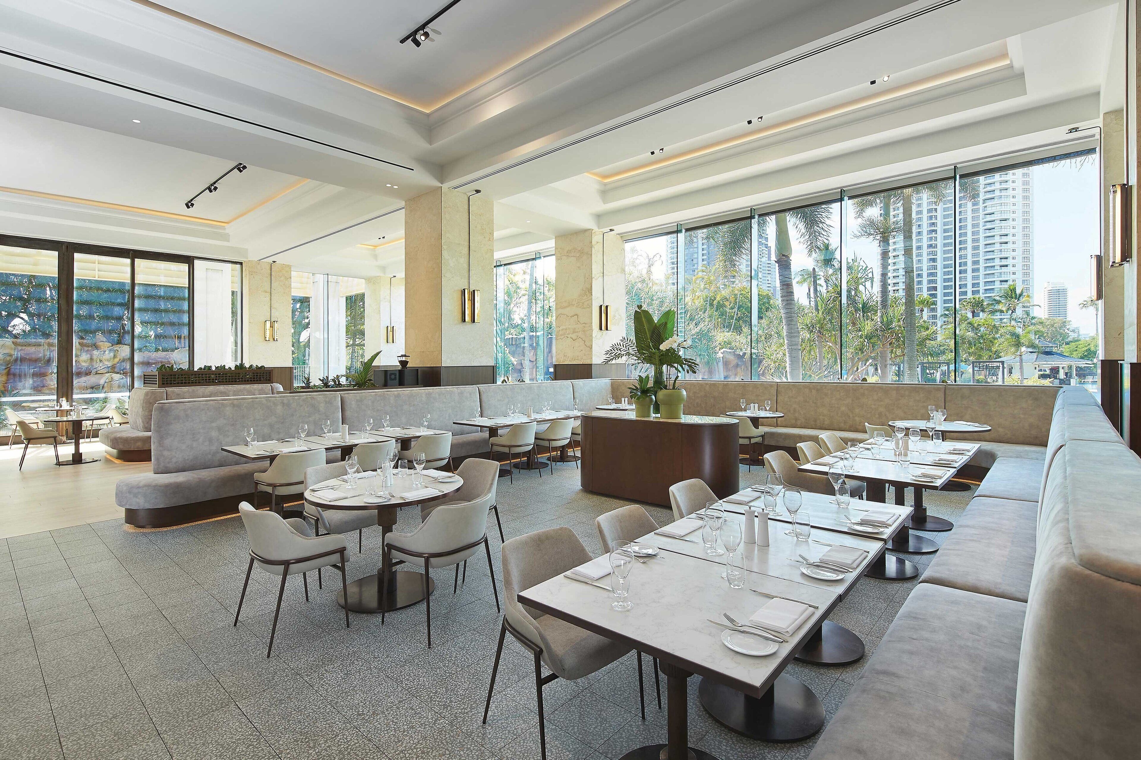 Um restaurante moderno com pé-direito alto, grandes janelas e vista para palmeiras e edifícios.