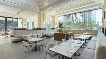 Um restaurante moderno com pé-direito alto, grandes janelas e vista para palmeiras e edifícios.