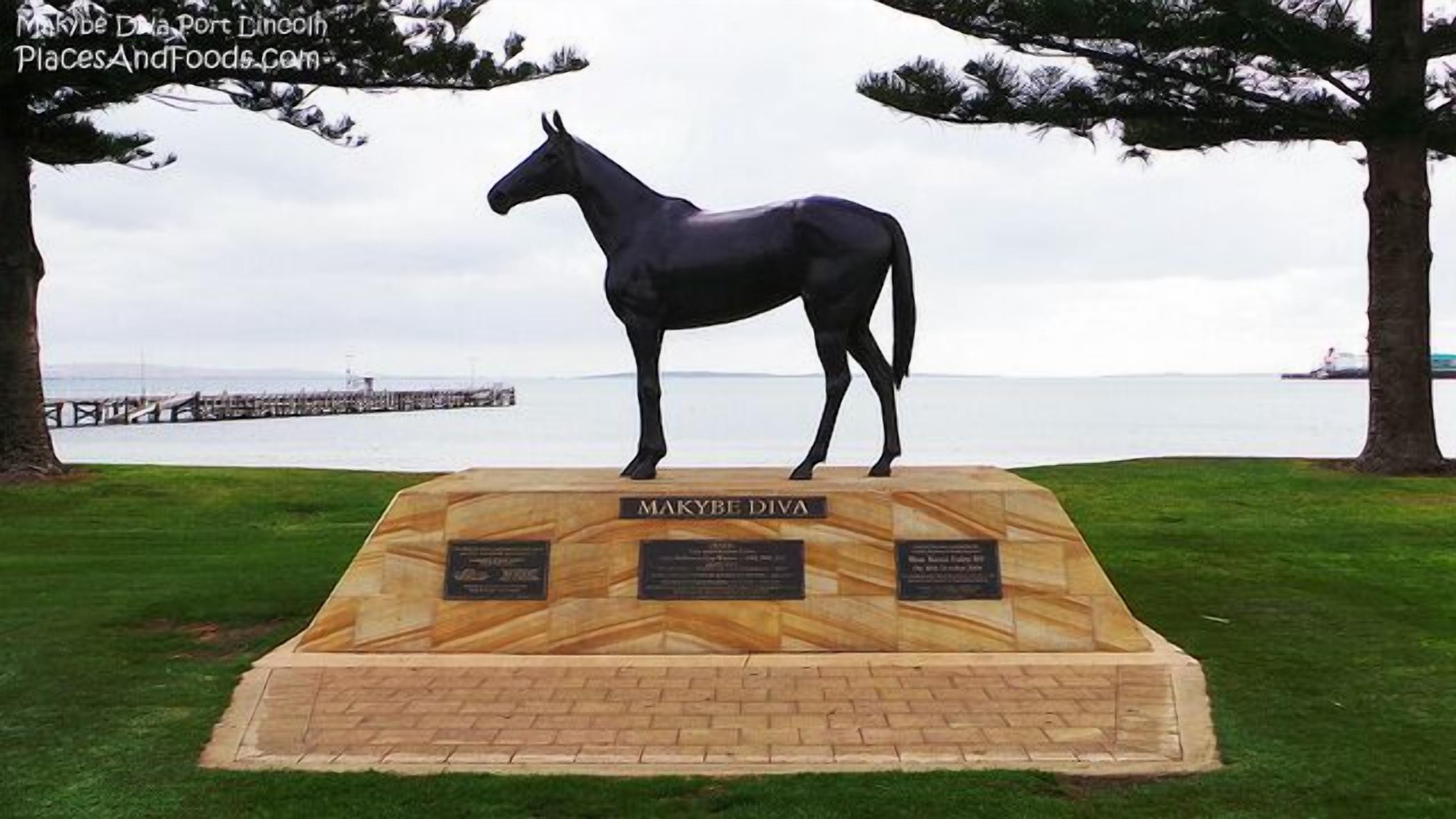 Uma estátua de cavalo em um pedestal com placas, com um corpo de água e árvores ao fundo.