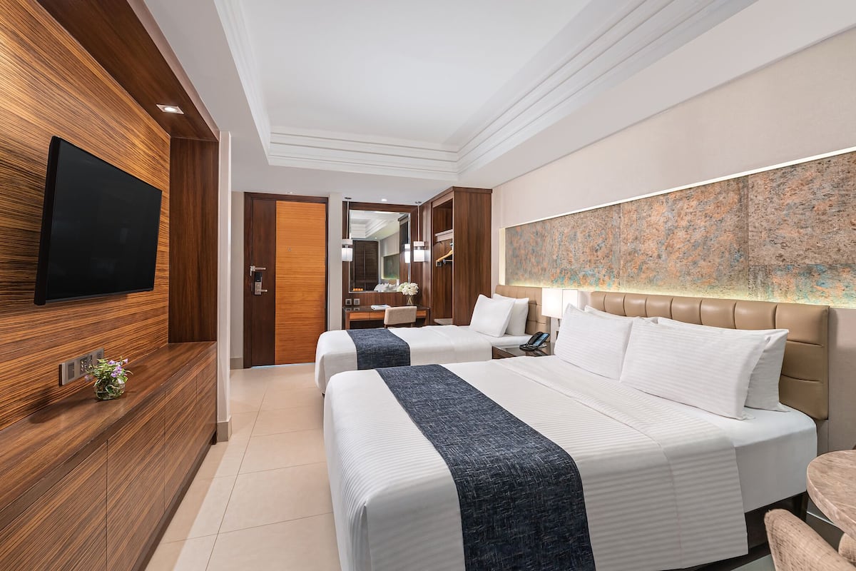 Quarto de hotel moderno com duas camas, uma TV de tela plana, um quadro abstrato grande e um espaço de refeições com uma travessa de frutas.