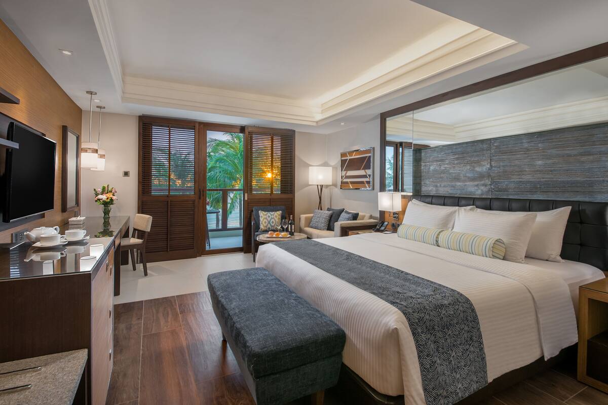 Quarto de hotel moderno com uma cama grande, uma escrivaninha com televisão, área de estar com vista e varanda com palmeiras.