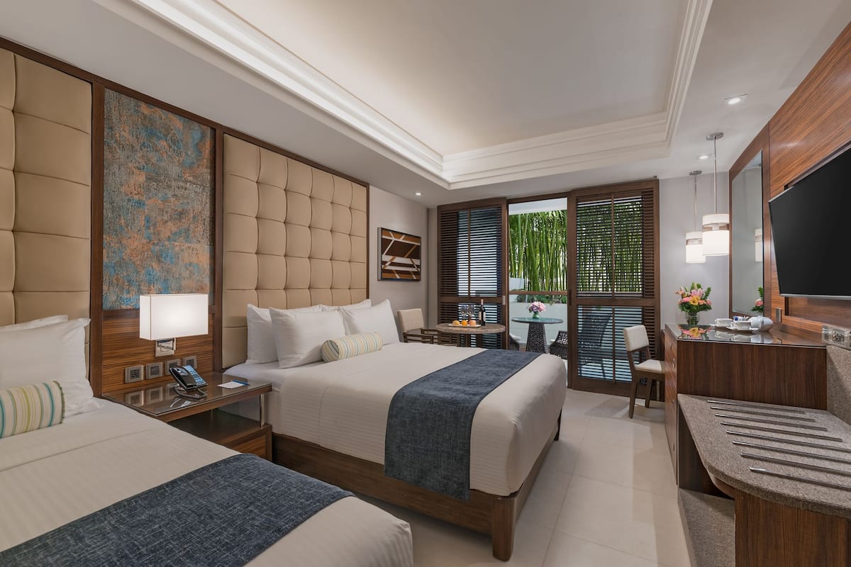 Quarto de hotel com duas camas, uma escrivaninha, televisão e vista para área verde.