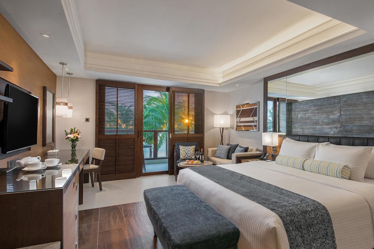 Quarto de hotel moderno com uma cama grande, escrivaninha com TV de tela plana, área de estar com sofá e poltronas, e varanda com vista para palmeiras.